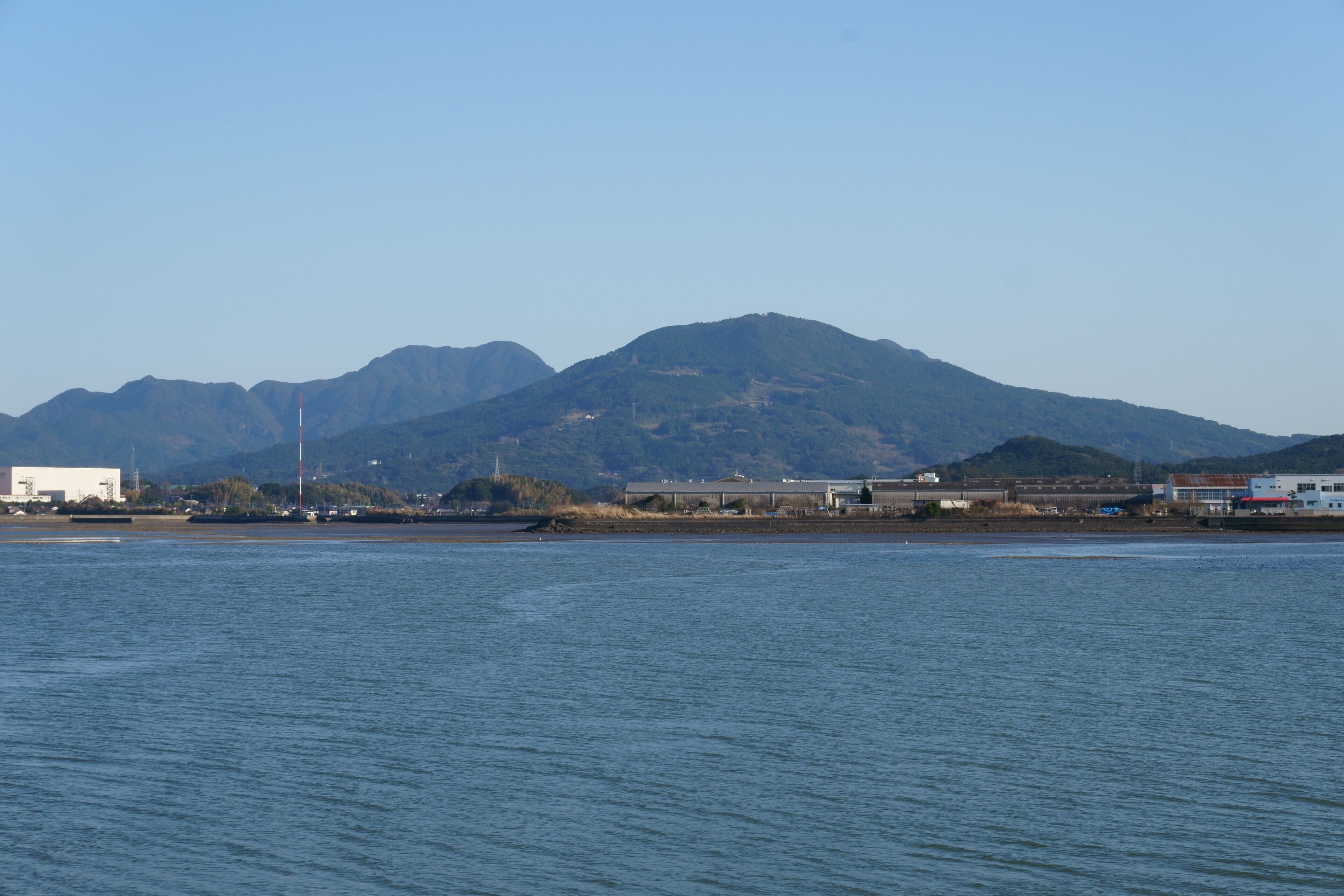 Água calma com montanhas ao fundo e edifícios industriais na margem.