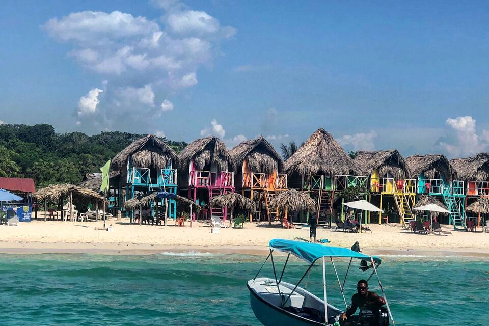 Um barco em águas turquesa com uma praia e edifícios coloridos ao fundo.