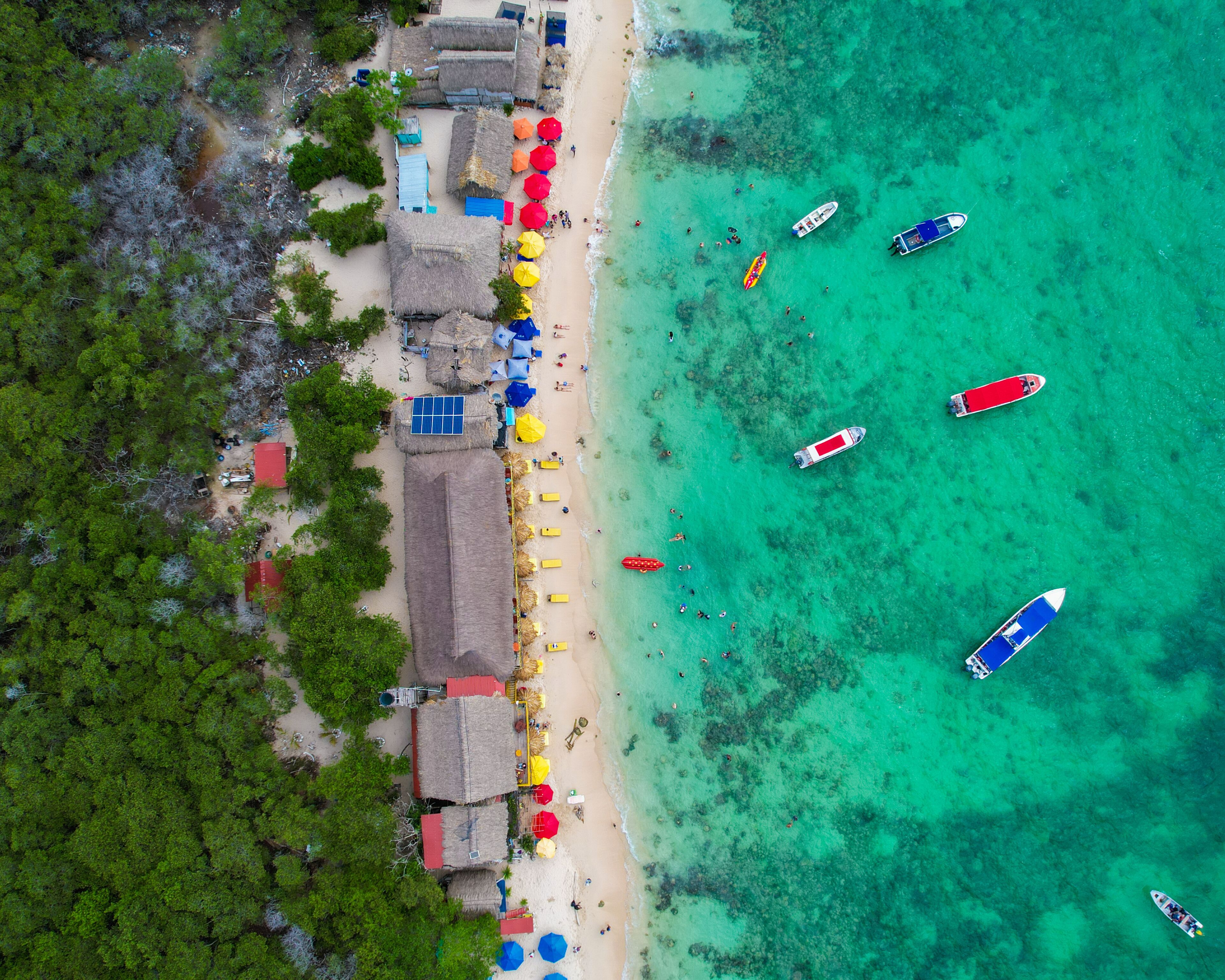 Vista aérea de uma praia com palafitas de telhado de palha, guarda-sóis coloridos e vários barcos ancorados em águas turquesa cristalinas.