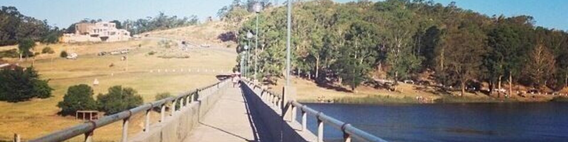 Ponte de concreto com grades de metal e luminária esférica, com vista para um rio e uma paisagem acidentada com vegetação escassa.