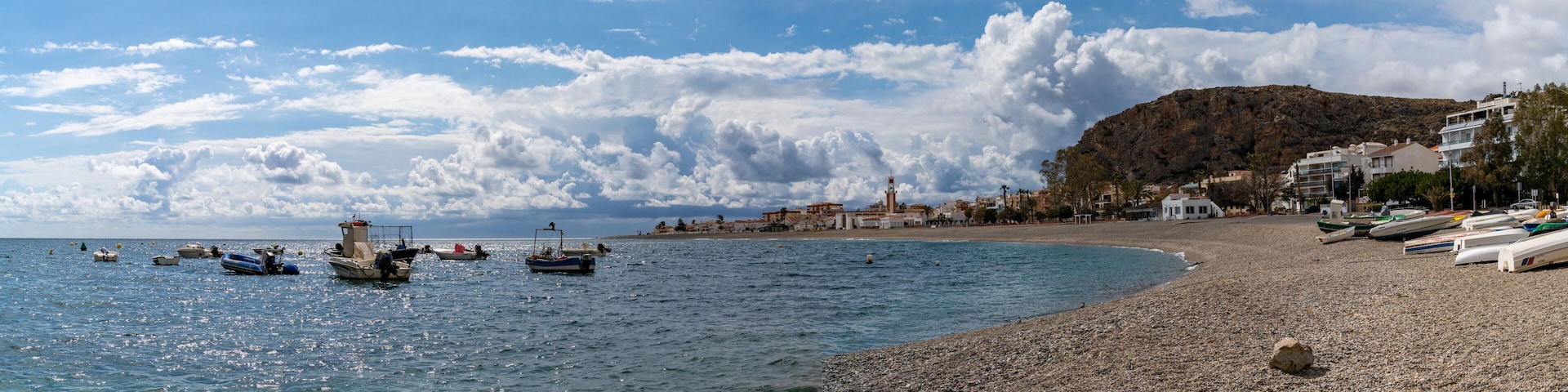 Uma praia de seixos com barcos, uma colina rochosa e edifícios ao fundo.