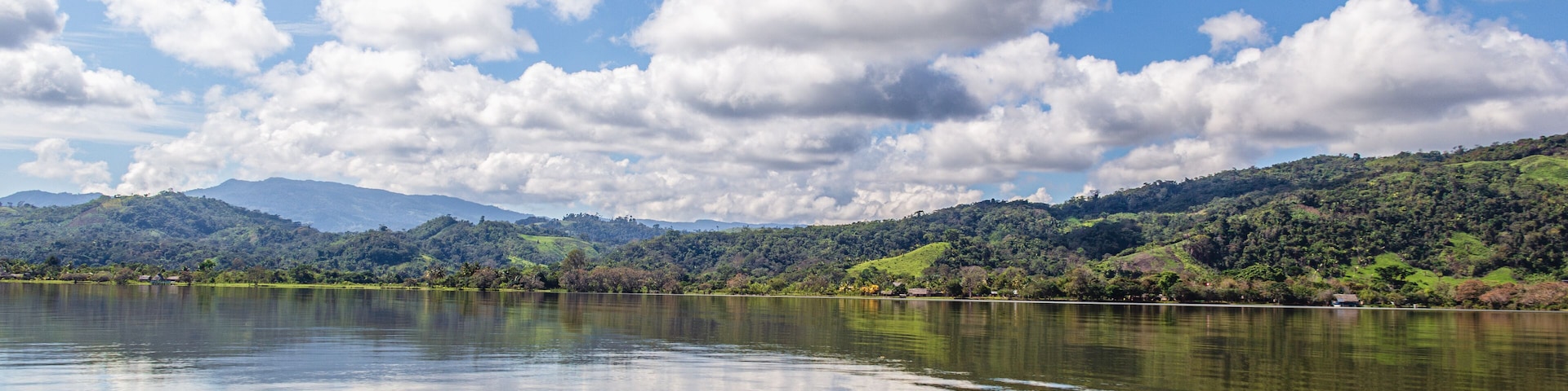 Um lago calmo com reflexos de nuvens, rodeado por colinas e montanhas verdes sob um céu azul límpido.