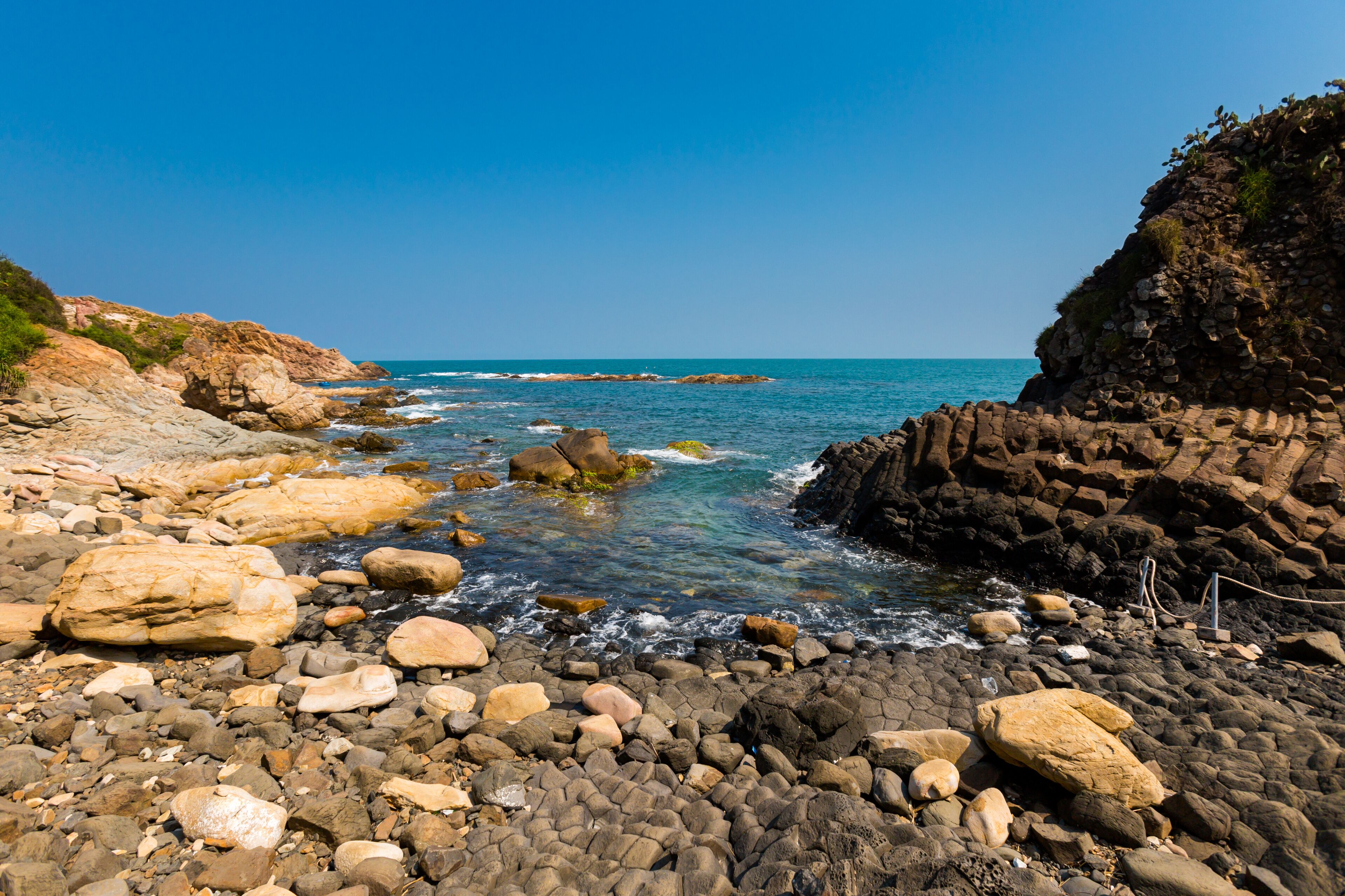 Litoral rochoso com águas azul-claras, pedras grandes e um penhasco íngreme.