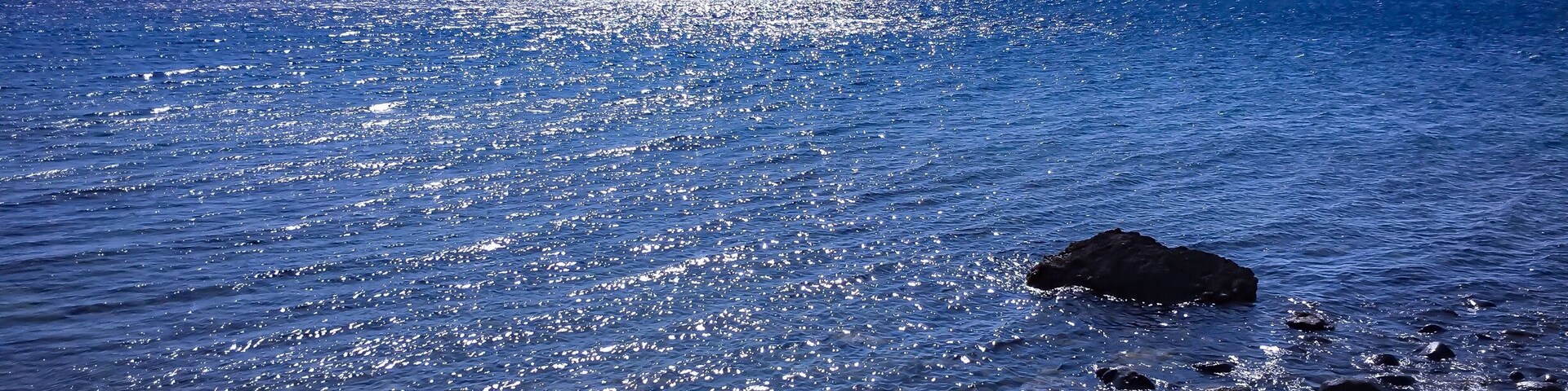 Uma praia com águas cristalinas refletindo a luz do sol, pedras espalhadas e ilhas ao longe sob um céu azul.