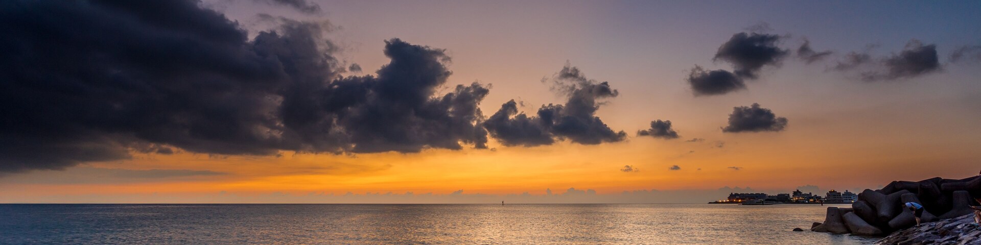 Um pôr do sol sobre um mar calmo, com costa rochosa e um horizonte com edifícios ao longe.