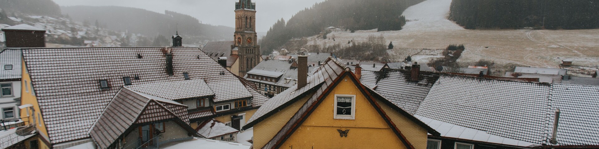 Uma aldeia coberta de neve com casas tradicionais, uma igreja e uma colina arborizada ao fundo.