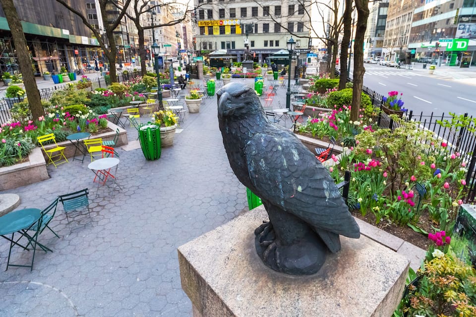 Escultura de um pássaro sobre um pedestal, rodeada por um jardim de flores e mesas ao ar livre.