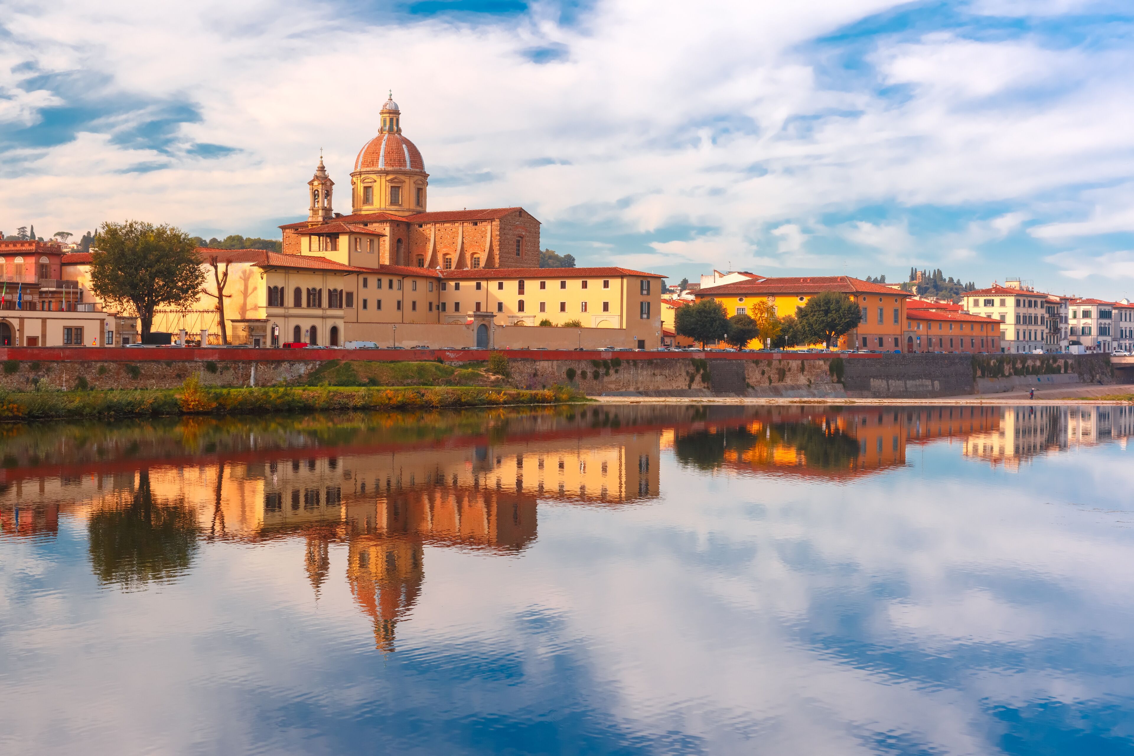 Um rio calmo refletindo edifícios históricos, incluindo uma igreja notável com cúpula e campanário.