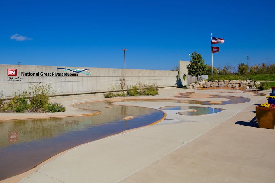 Entrada do Museu Nacional dos Grandes Rios, com um muro de concreto, bandeiras americanas e uma área ajardinada com elementos aquáticos.