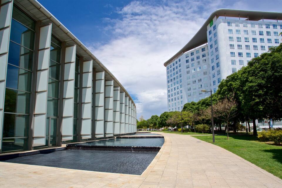 Um edifício moderno com grandes janelas e um telhado curvo, rodeado por um parque bem cuidado, com árvores e um caminho pavimentado.