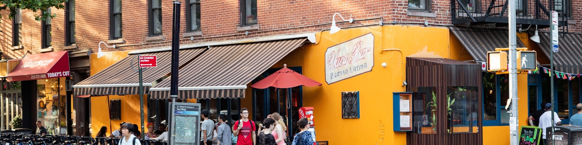 Um movimento esquina de rua com um prédio amarelo, toldo vermelho e uma placa com o nome 'Pura Vida Cocina Latina'.