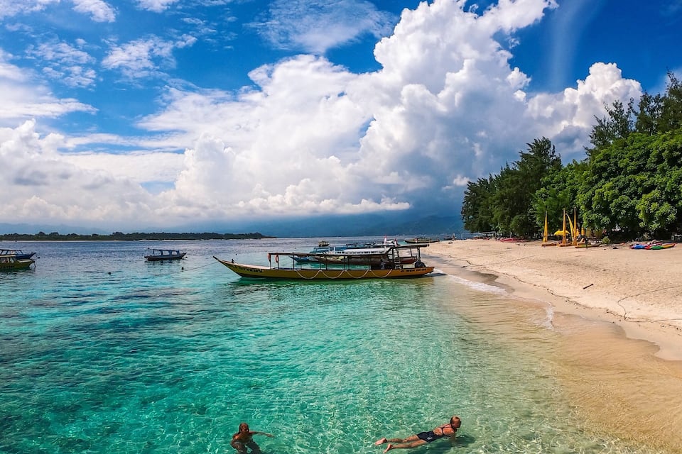 Uma praia com águas turquesa cristalinas, algumas pessoas nadando e barcos tradicionais ancorados perto da costa.