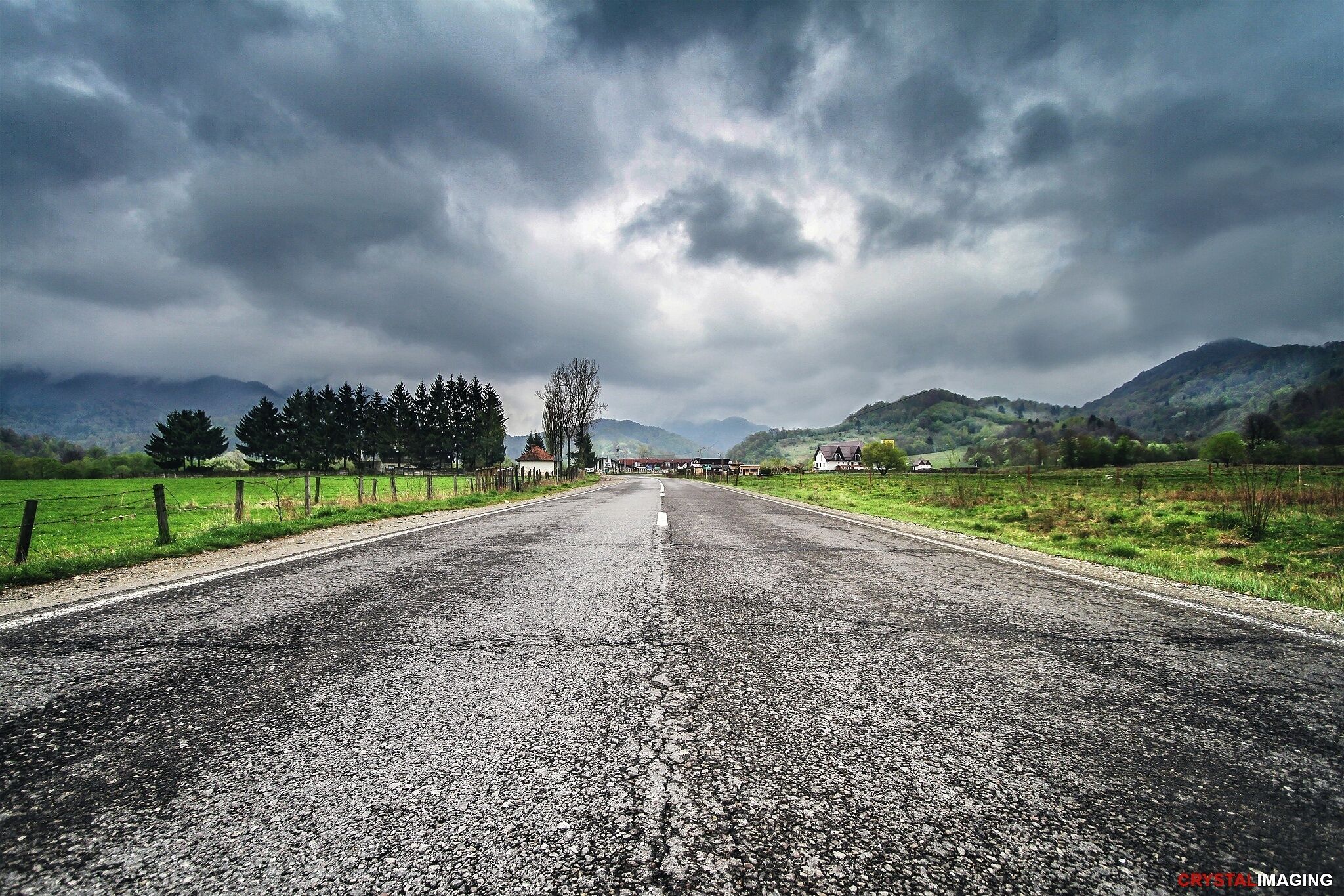Uma estrada rural com cerca à esquerda, uma árvore ao centro e montanhas ao fundo.
