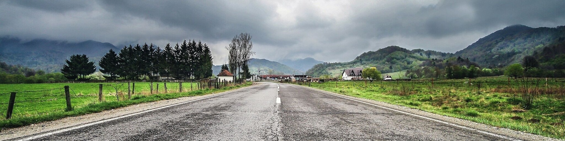 Uma estrada rural com cerca à esquerda, uma árvore ao centro e montanhas ao fundo.