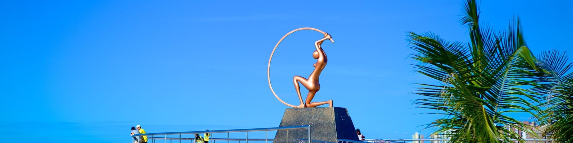 Praia com escultura de figura humana segurando um aro, palmeiras e céu azul límpido.