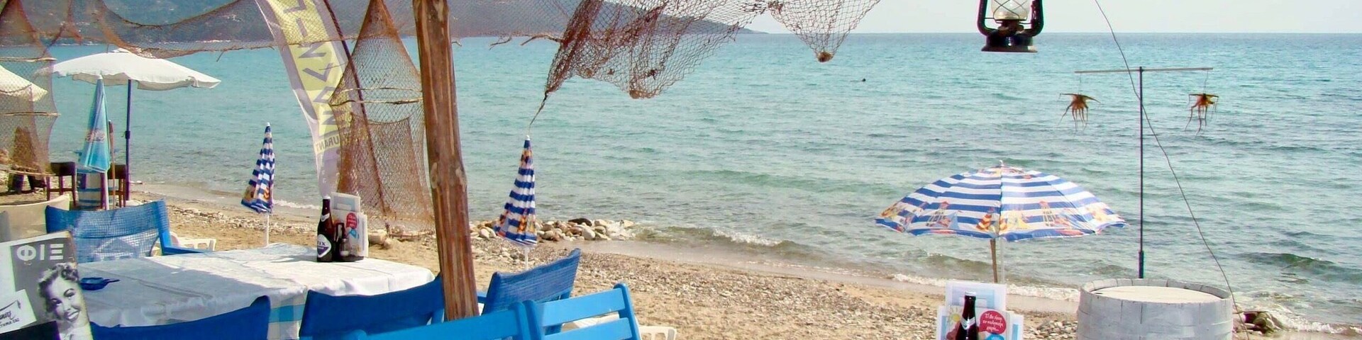 Um café à beira-mar com cadeiras e mesas azuis, um toldo de palha e uma área de pesca com redes.