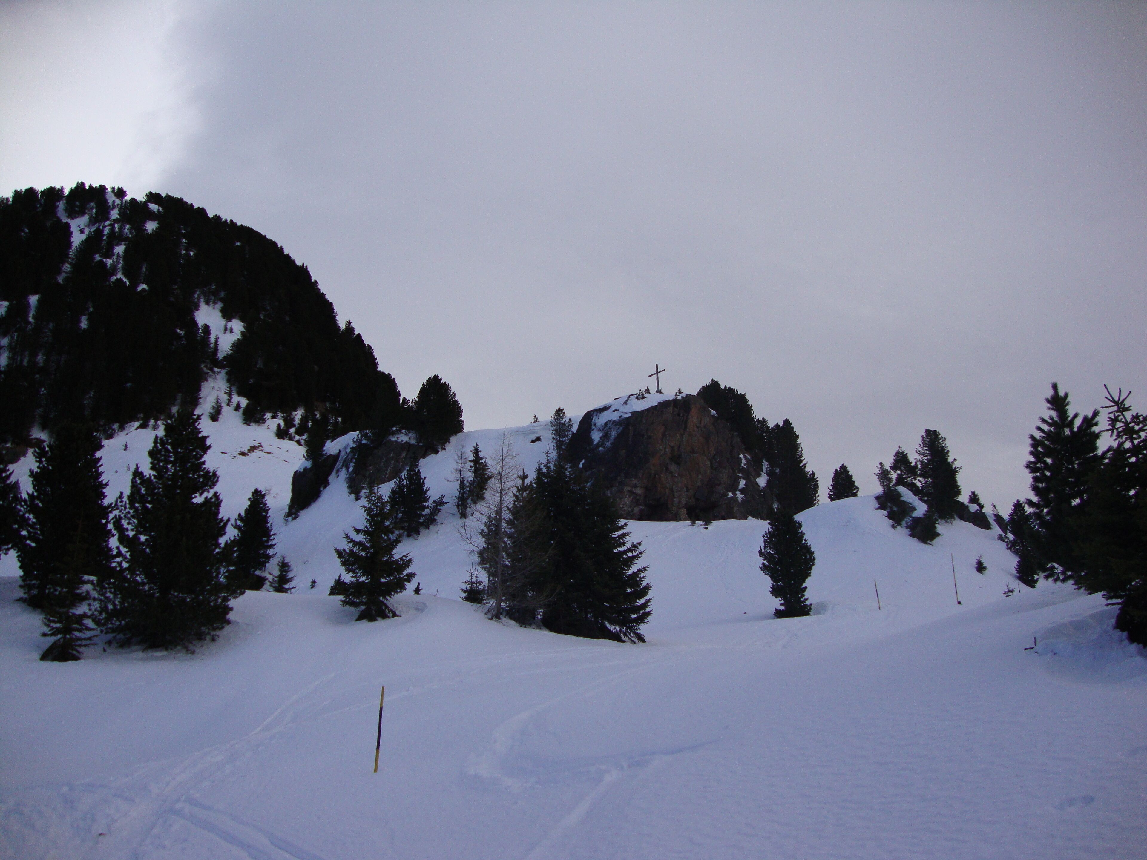 Montanha coberta de neve com uma cruz no topo, cercada por árvores perenes.