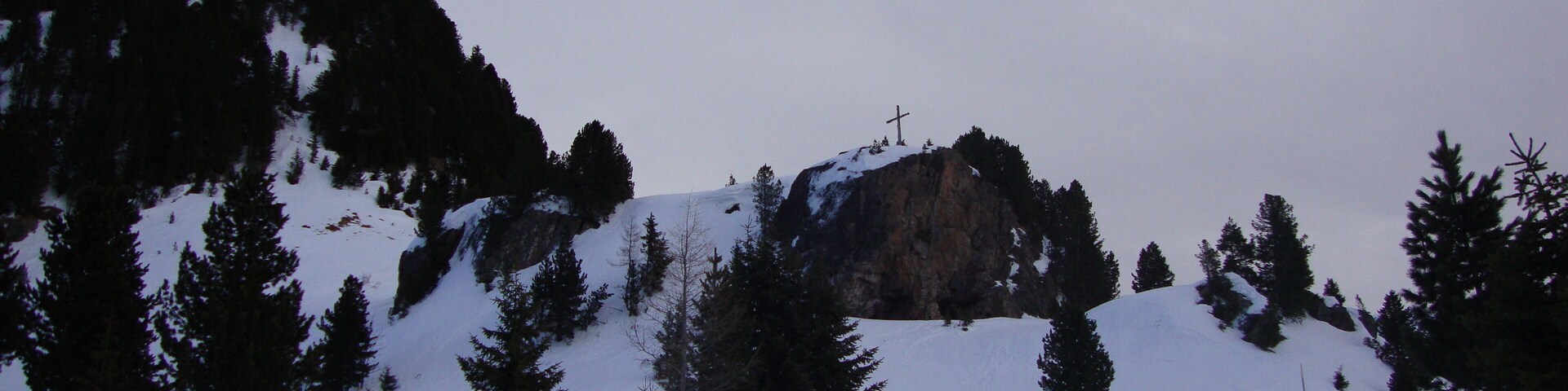 Montanha coberta de neve com uma cruz no topo, cercada por árvores perenes.