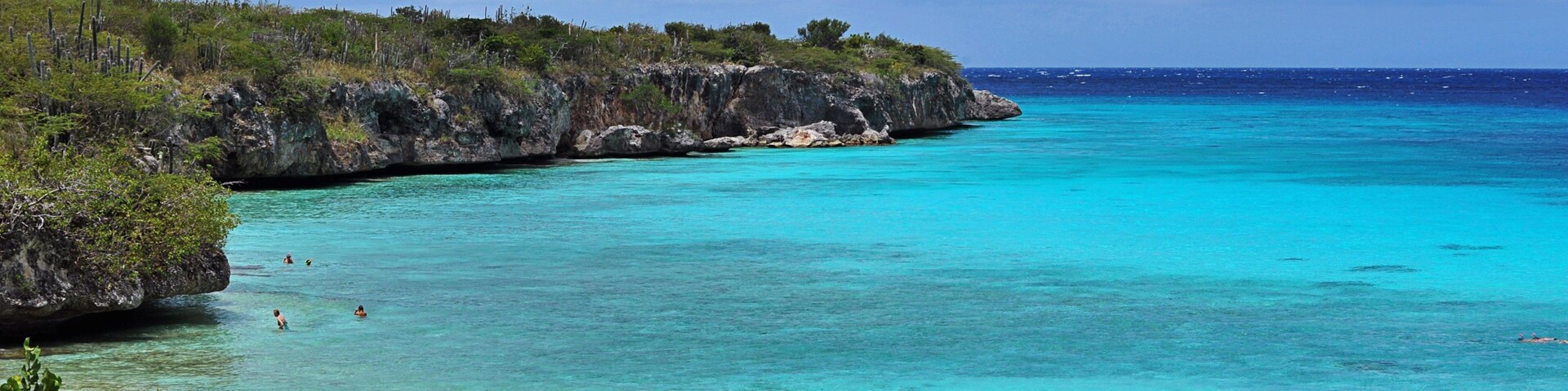 Mar azul-turquesa, falésias rochosas e um cais de madeira com guarda-sóis.