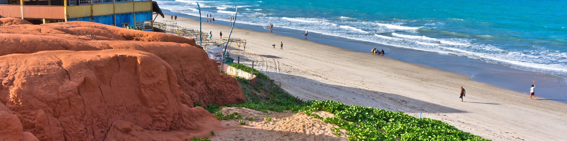 Uma praia com areia vermelha, vegetação verde e um edifício com telhado azul.