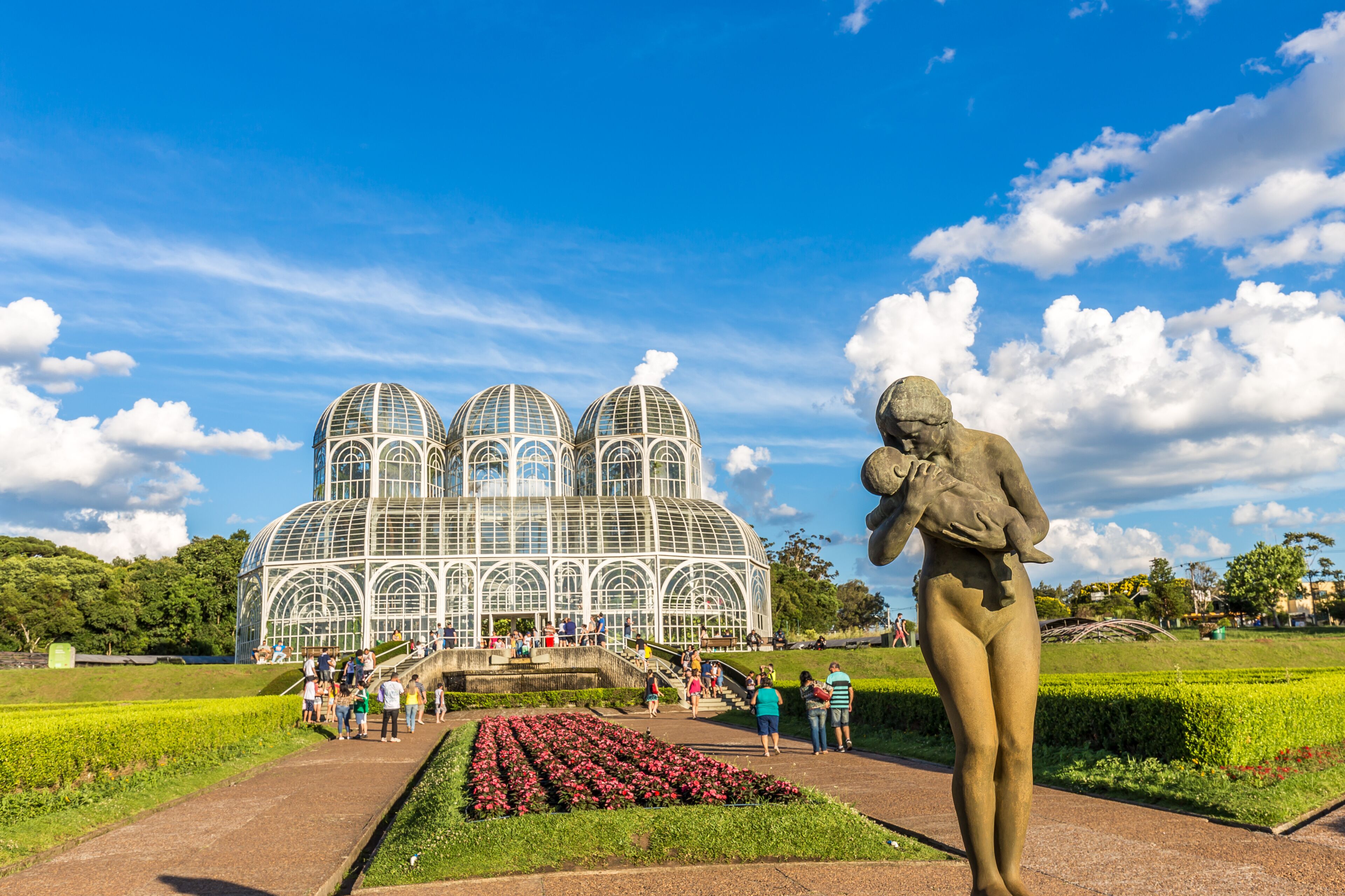 Uma grande estufa de vidro com várias cúpulas, rodeada por um jardim bem cuidado e uma estátua de uma figura segurando uma criança.