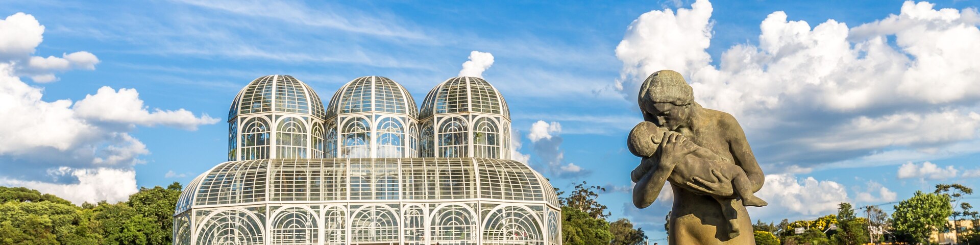 Uma grande estufa de vidro com várias cúpulas, rodeada por um jardim bem cuidado e uma estátua de uma figura segurando uma criança.