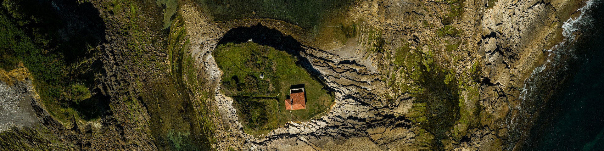 Vista aérea de uma área costeira com um pequeno edifício rodeado por terreno rochoso e vegetação verde.