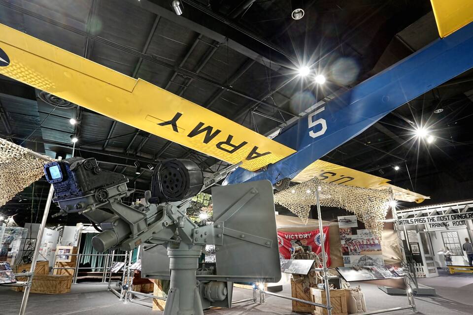 Exposição de aeronaves militares com uma metralhadora montada e diversos painéis informativos.