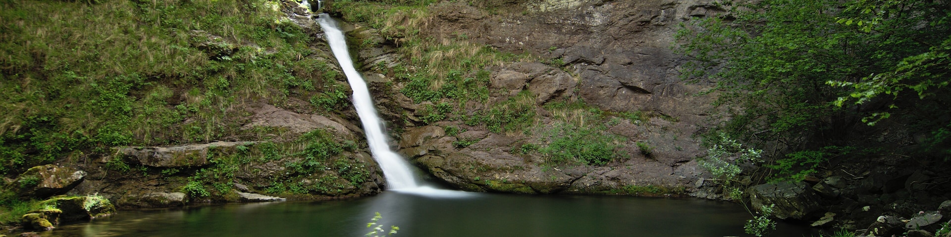 Uma cascata deságua em uma piscina límpida, rodeada por rochas e vegetação.