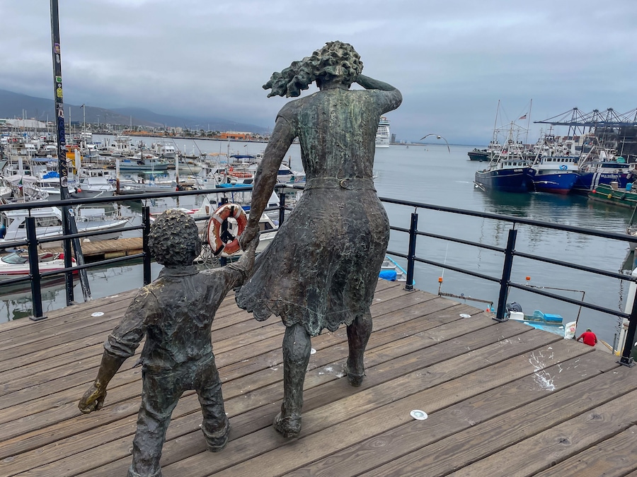 Uma estátua de uma mulher e uma criança com vista para uma marina com barcos e uma ponte ao longe.