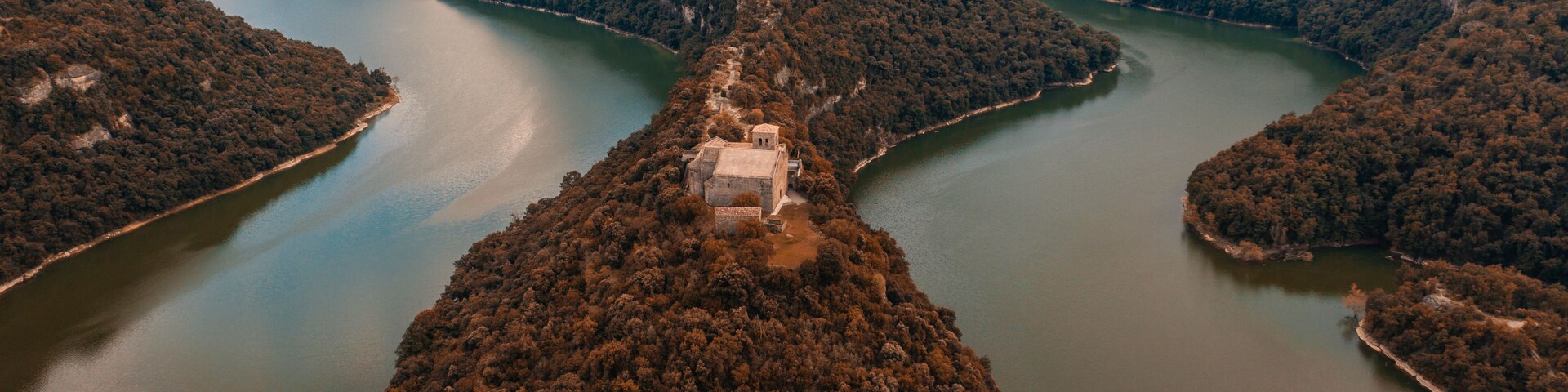 Vista aérea de uma ilha arborizada com um pequeno edifício, cercada por um rio e colinas.