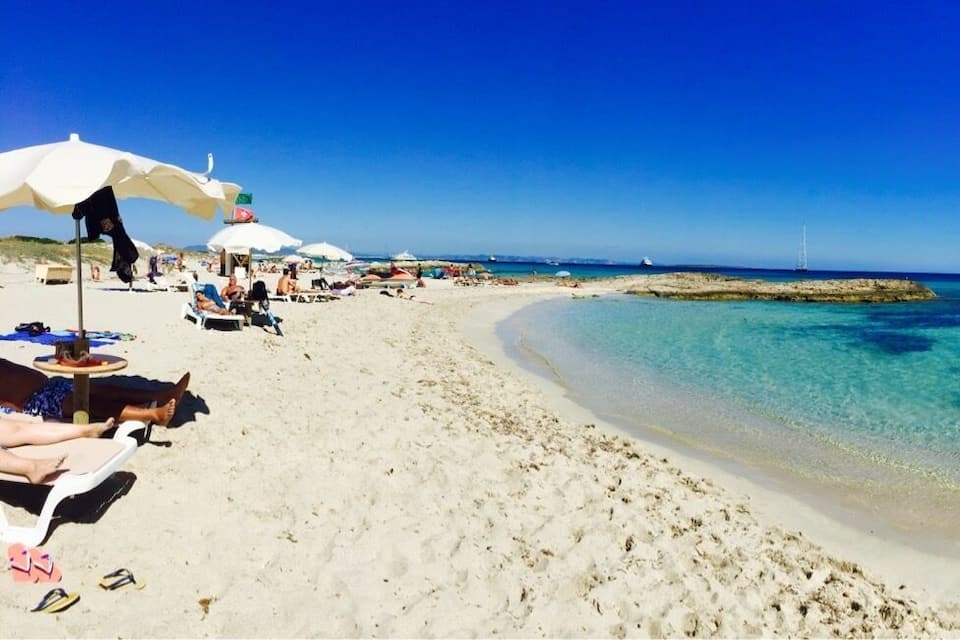 Uma praia com águas turquesa cristalinas, areia dourada e algumas pessoas aproveitando o sol.