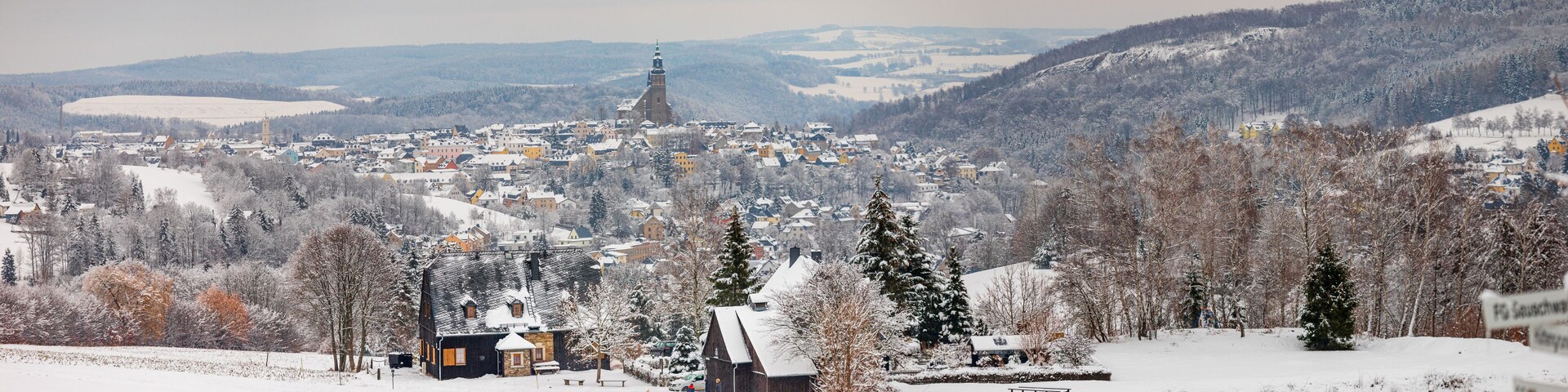 Uma cidade coberta de neve com uma torre de igreja imponente, cercada por colinas arborizadas.