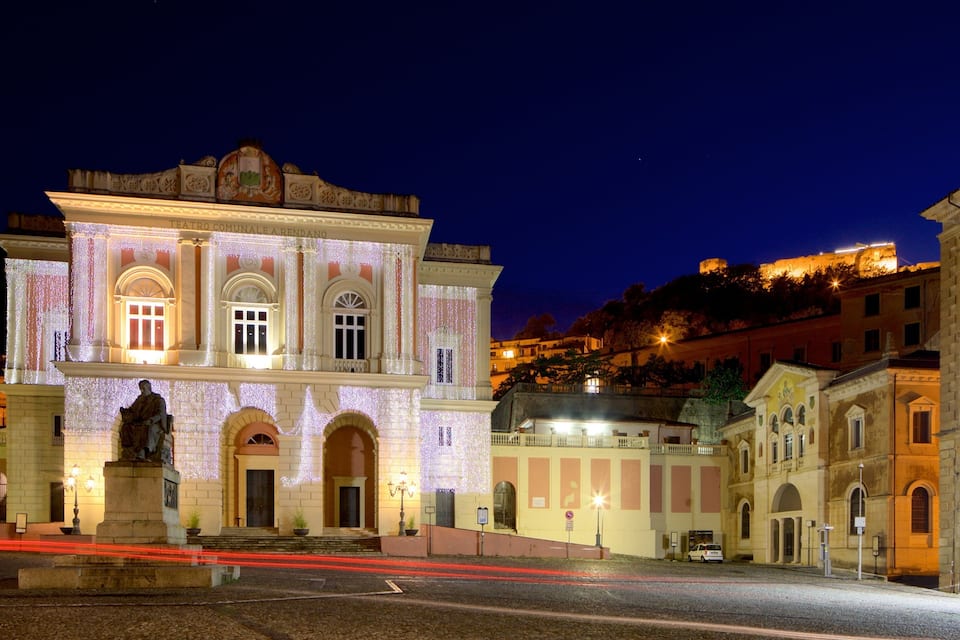 Teatro Comunale A. Rendano com arcos iluminados e uma estátua em frente.