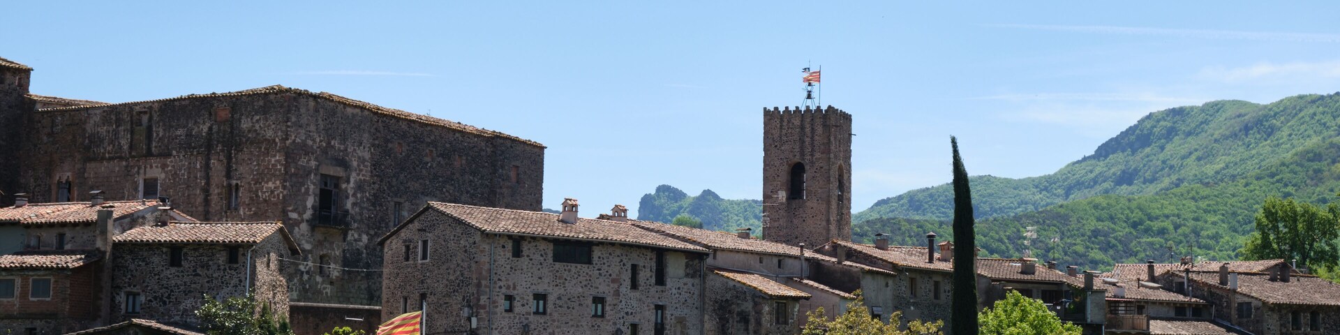 Uma cidade histórica com edifícios de pedra, uma torre imponente e colinas verdejantes ao fundo.