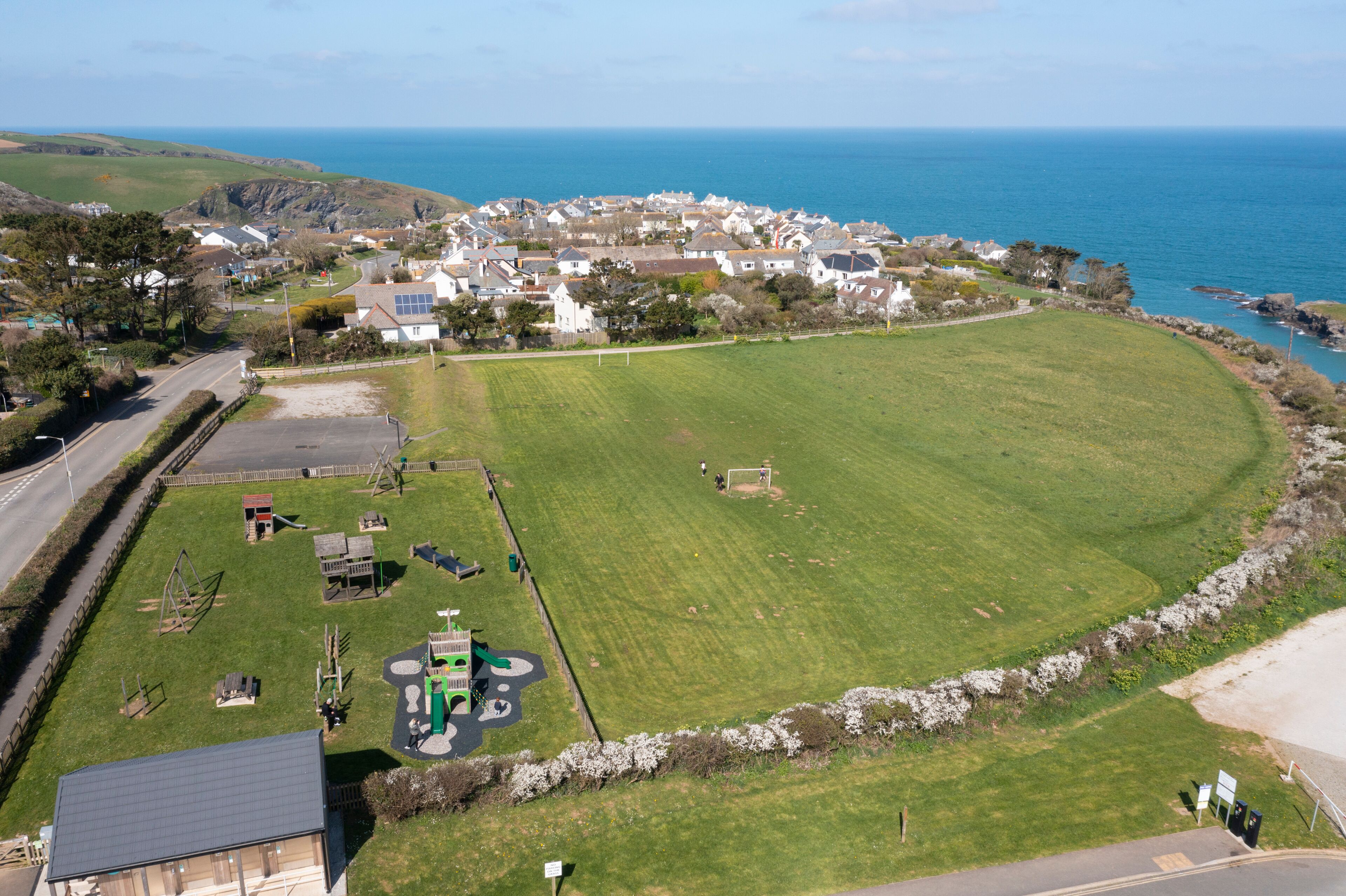 Uma vila costeira com um vasto campo verde, um parquinho e casas perto do mar.