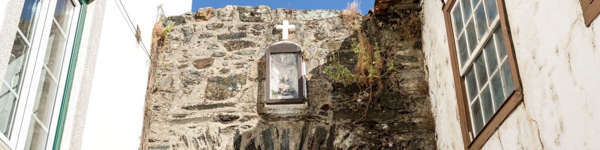 Um arco de pedra com uma cruz e um quadro emoldurado acima, cercado por edifícios com fachadas brancas e marrons.