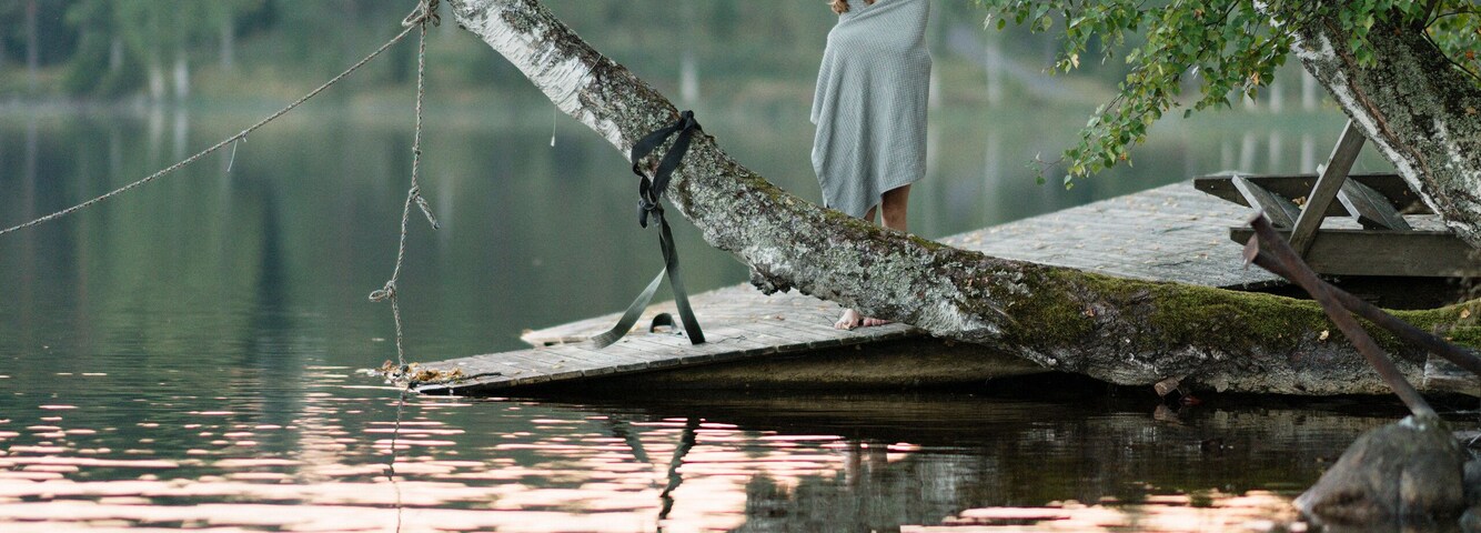 Uma mulher está em um píer à beira de um lago calmo, cercada por árvores.