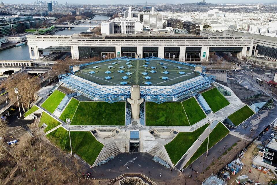 Um edifício grande e moderno com uma estrutura de telhado circular única, rodeado por relvados verdes e um rio ao fundo.
