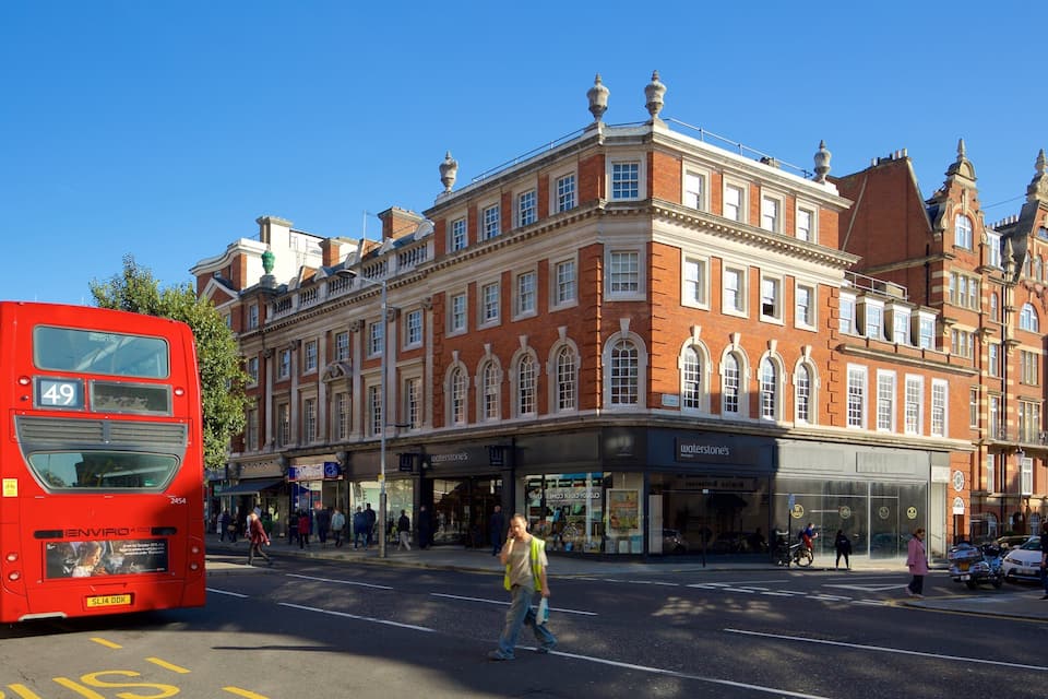 Um ônibus de dois andares vermelho em uma rua da cidade, um edifício histórico com janelas em arco e pedestres.