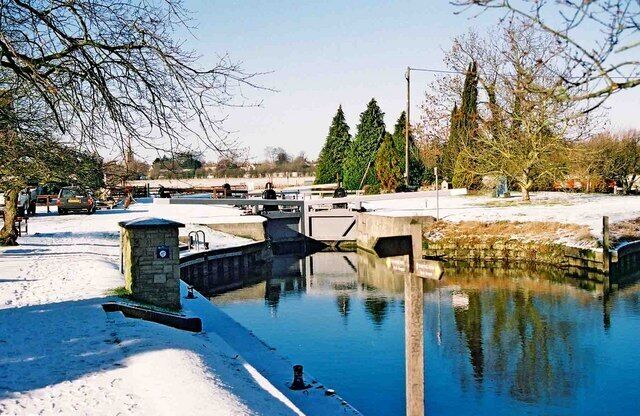 Um canal coberto de neve, com uma ponte de pedra e árvores ao fundo.