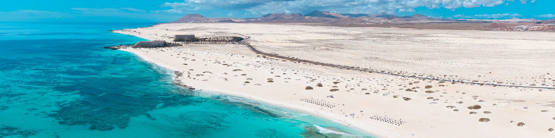 Paisagem costeira com praia arenosa, águas turquesas cristalinas e montanhas ao longe sob um céu azul.