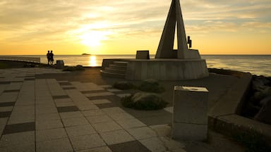 Um pôr do sol sobre um calçadão costeiro, com um monumento e duas pessoas caminhando.