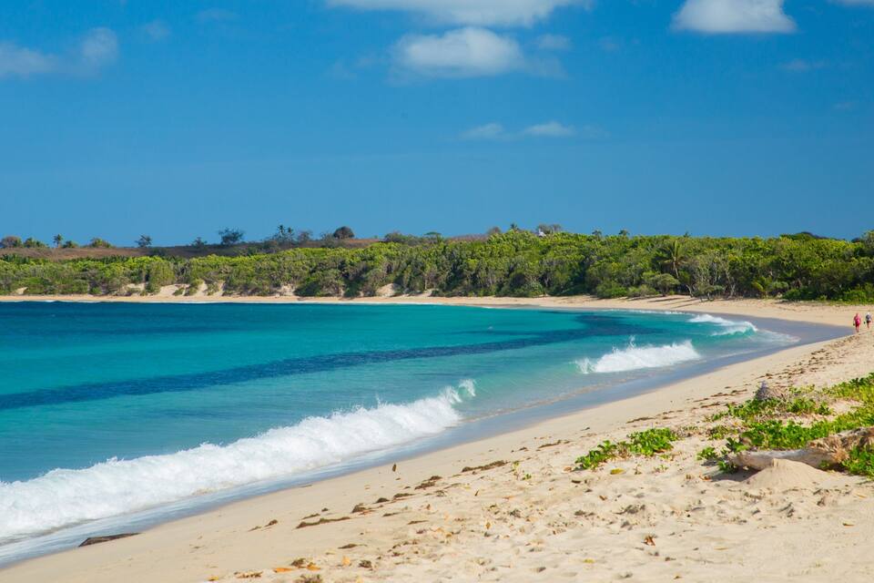 Uma praia com águas turquesa cristalinas, areal arenoso e vegetação verde.
