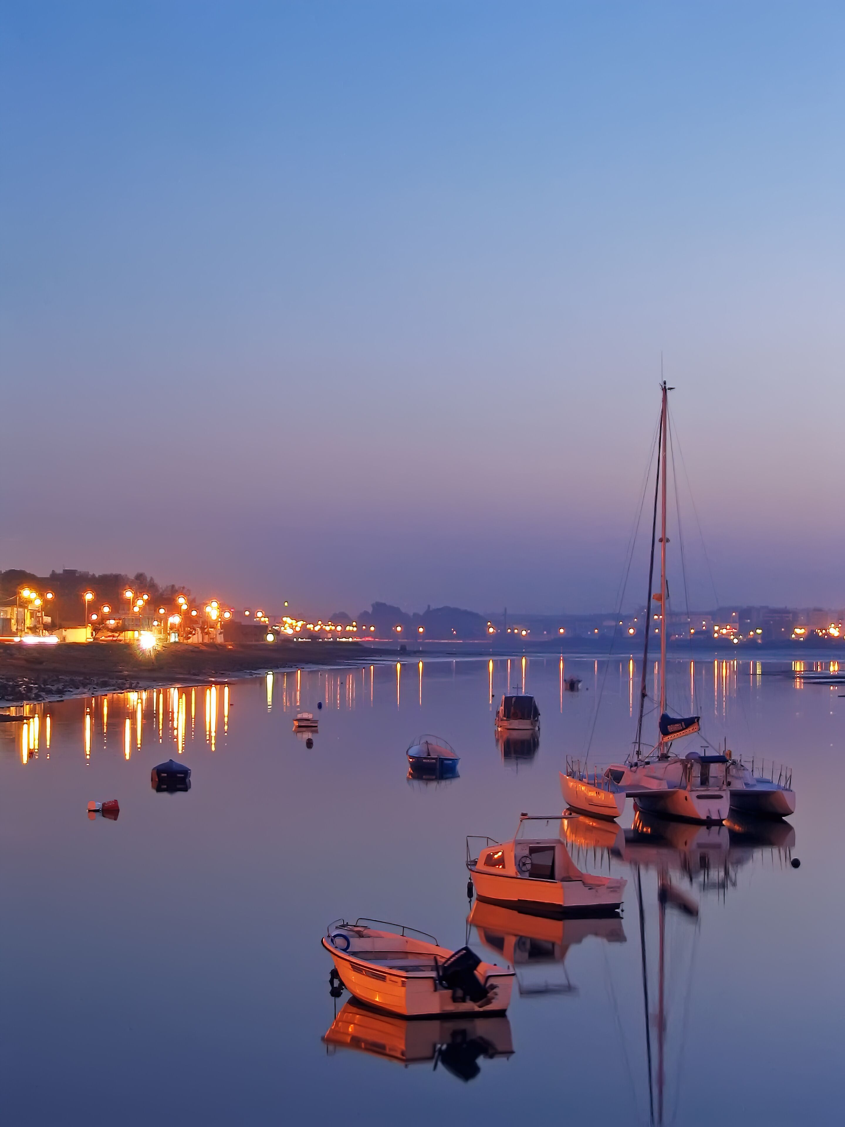 Um porto calmo com várias embarcações ancoradas, céu claro e paisagem urbana com luzes ao fundo.
