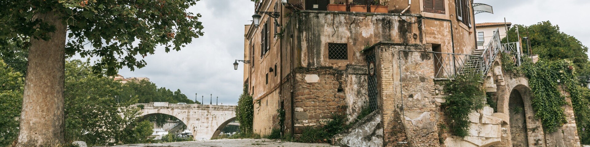 Um edifício histórico com uma escadaria de pedra que o leva até à entrada, rodeado por árvores e uma ponte de pedra ao fundo.