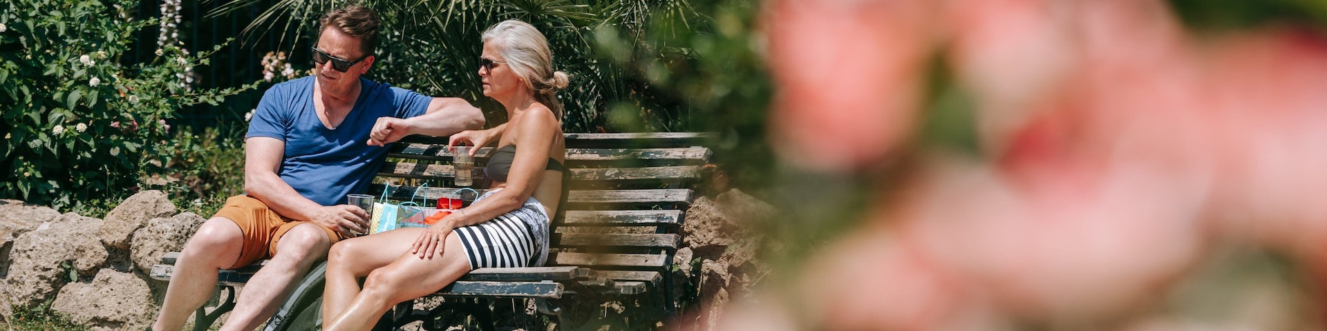 Um casal sentado em um banco de madeira, cercado por vegetação e pedras.