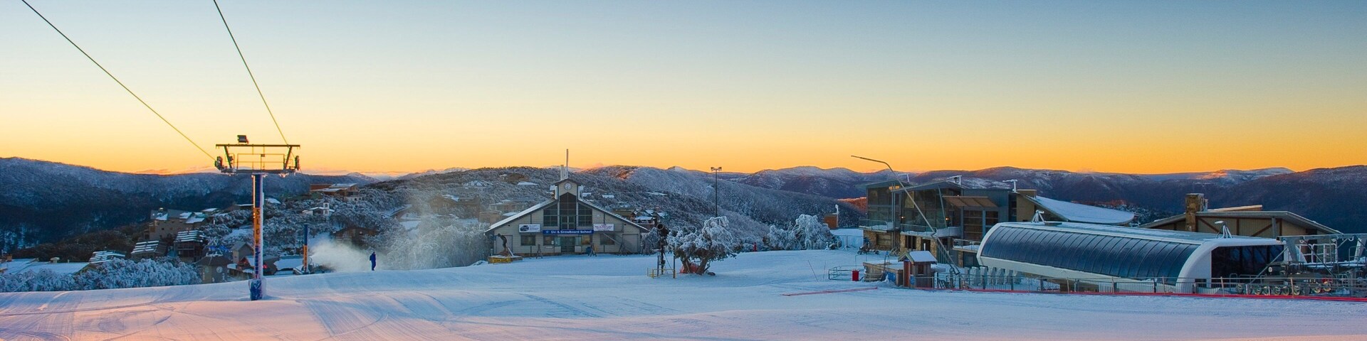 Pista de esqui coberta de neve, com teleférico e edifícios ao pôr do sol.