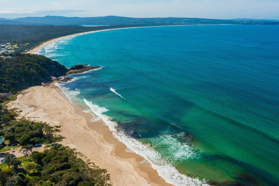 Paisagem costeira com uma praia arenosa, águas turquesa cristalinas e uma área arborizada.