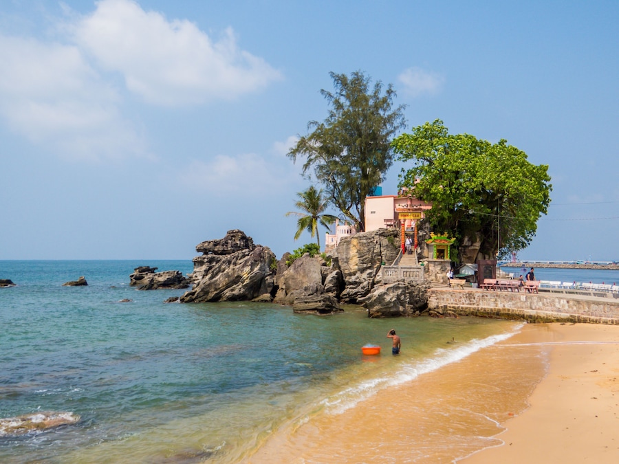 Uma praia com águas turquesa cristalinas, afloramentos rochosos, uma palmeira e um edifício com fachada colorida.